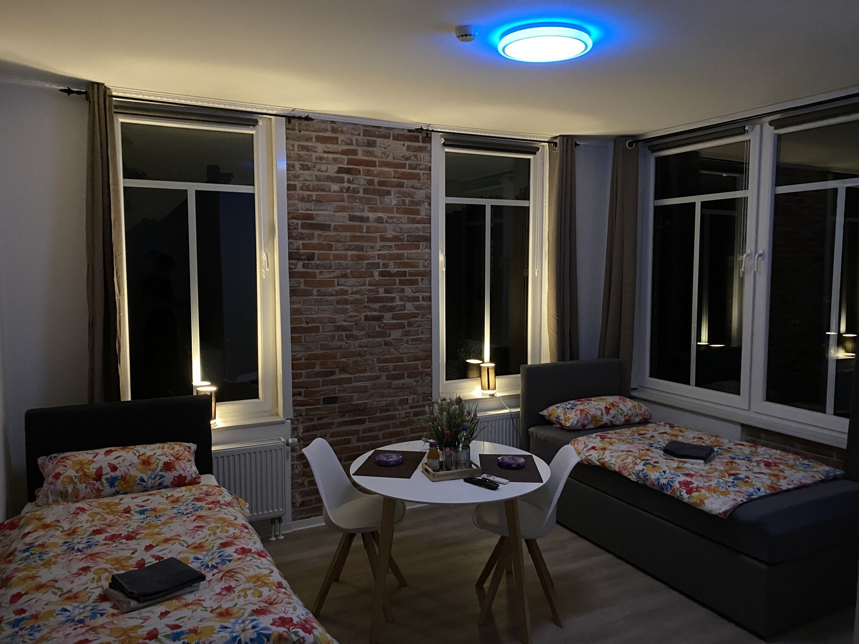 Quarto com duas camas, uma mesa e cadeiras, janelas amplas e parede de tijolos.
