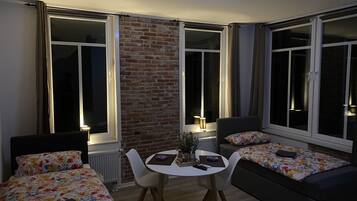 Quarto com duas camas, uma mesa e cadeiras, janelas amplas e parede de tijolos.