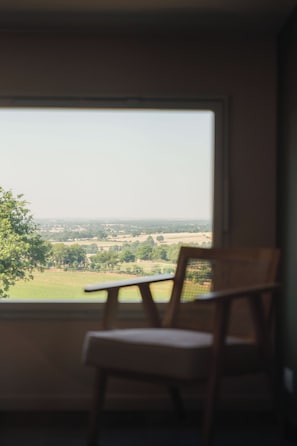 Quarto com uma única cadeira de madeira virada para uma janela ampla com vista para uma paisagem rural.