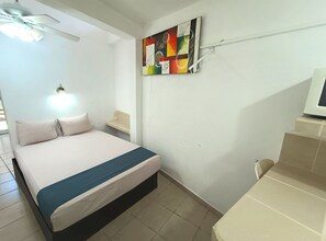 Quarto de hotel com cama, ventoinha no teto, televisão fixada na parede e uma pequena mesa.