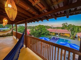 Varanda de madeira com uma rede, piscina com guarda-sóis e vista para um edifício com telhado vermelho.