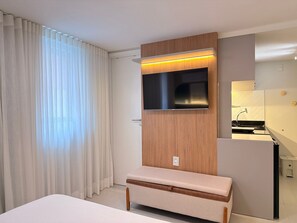 Quarto de hotel com cama, um banquinho, uma TV presa a um painel de madeira, espelho sobre a pia e janela com cortinas.