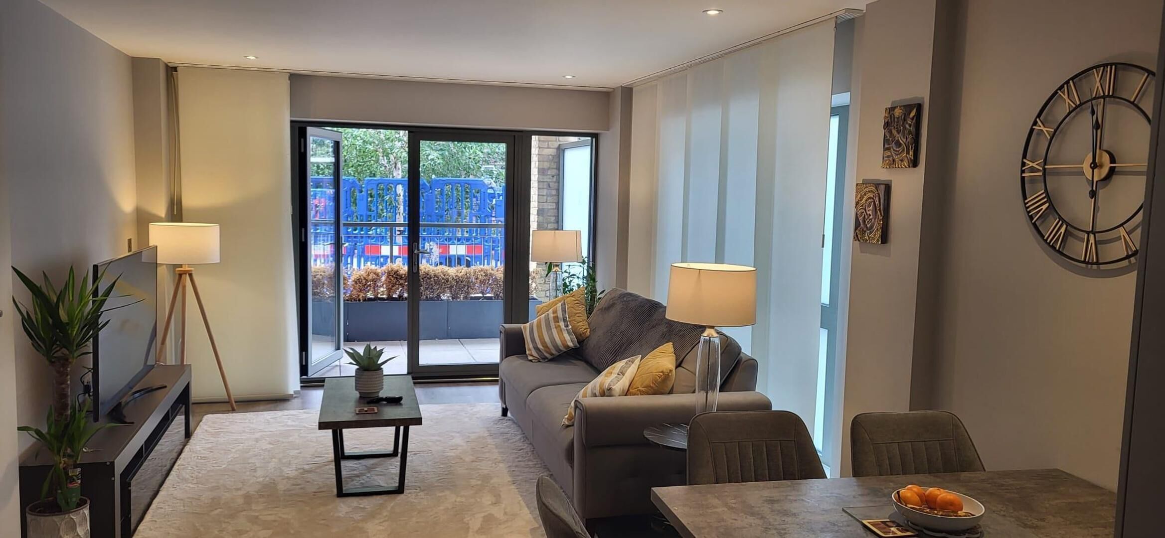 Uma sala de estar moderna com sofá, mesa de centro e mesa de jantar. Há uma janela grande com vista para uma construção azul lá fora.