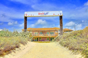 Um hotel à beira-mar com um letreiro indicando 'BeachGate CondoSuites & Hotel', um caminho de areia que leva à entrada e um edifício com varandas.
