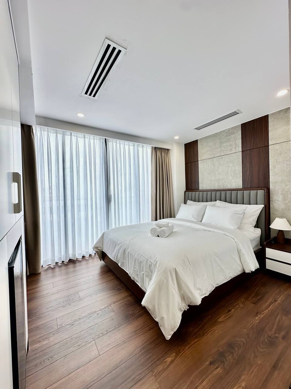 Quarto de hotel moderno com uma cama grande, piso de madeira, painel de cabeceira embutido e uma janela com cortinas.