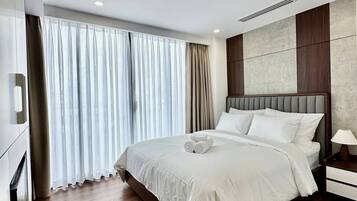 Quarto de hotel moderno com uma cama grande, piso de madeira, painel de cabeceira embutido e uma janela com cortinas.