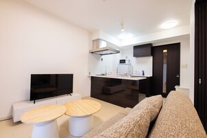 Uma sala de estar moderna com televisão de tela plana, duas mesas redondas de madeira, um sofá bege, área de cozinha com micro-ondas e uma porta de madeira escura.