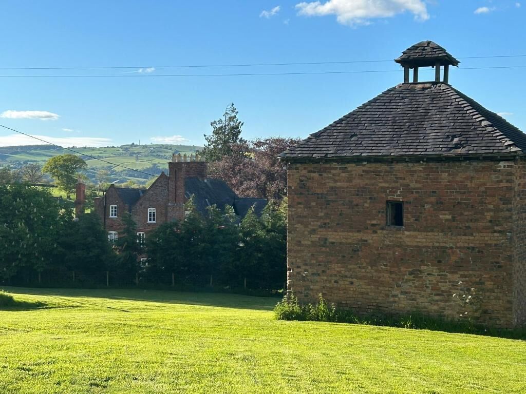 Um edifício de tijolos com telhado inclinado e uma pequena estrutura semelhante a uma torre no topo, cercado por um campo gramado e árvores.