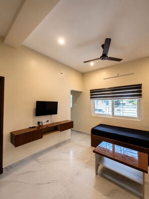 Uma sala de estar moderna com televisão de tela plana, estantes de madeira embutidas, um banco, um ventilador de teto e uma janela com persianas.