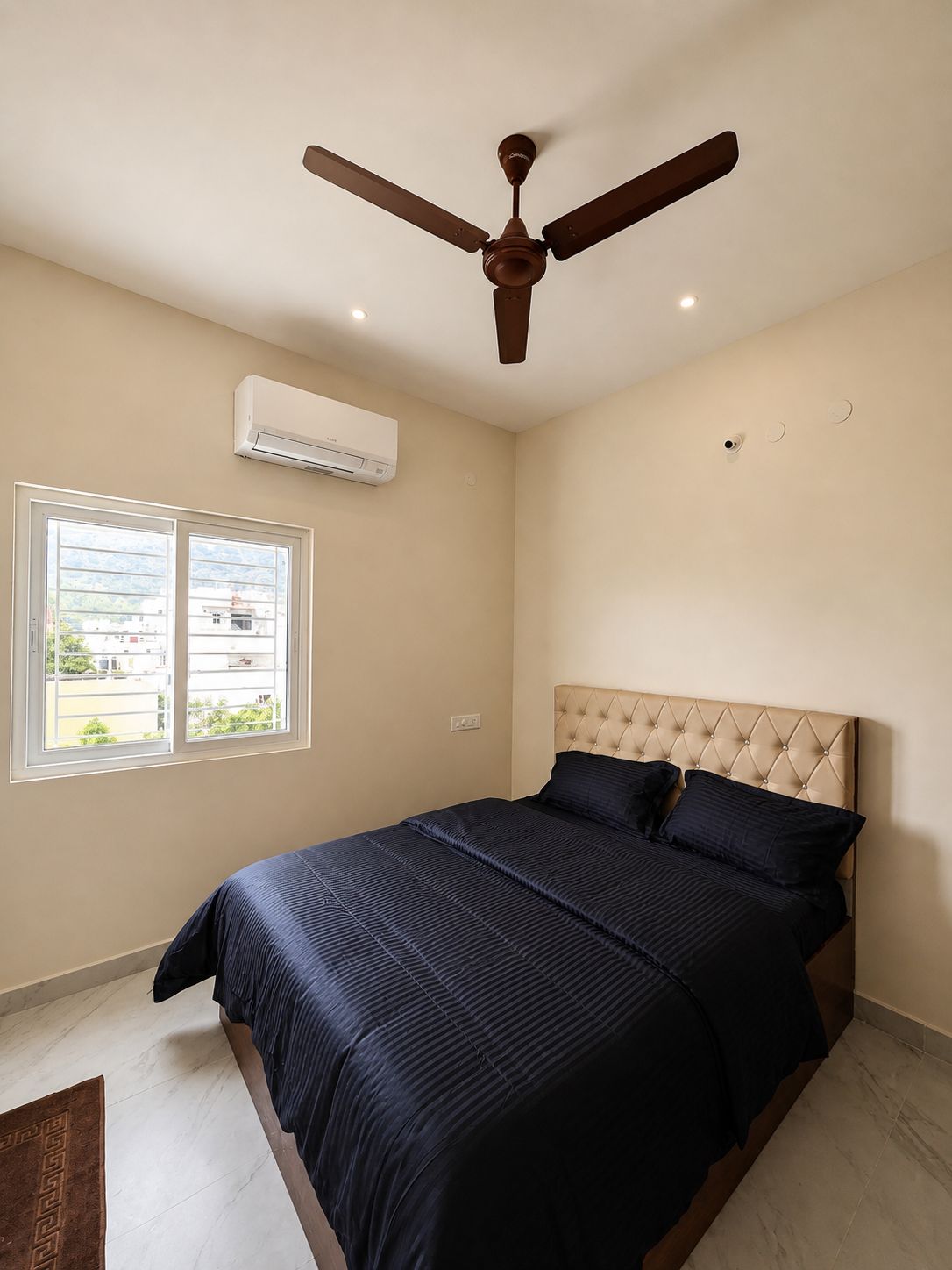 Quarto com cama, ventilador de teto, ar condicionado e janela com vista para edifícios.