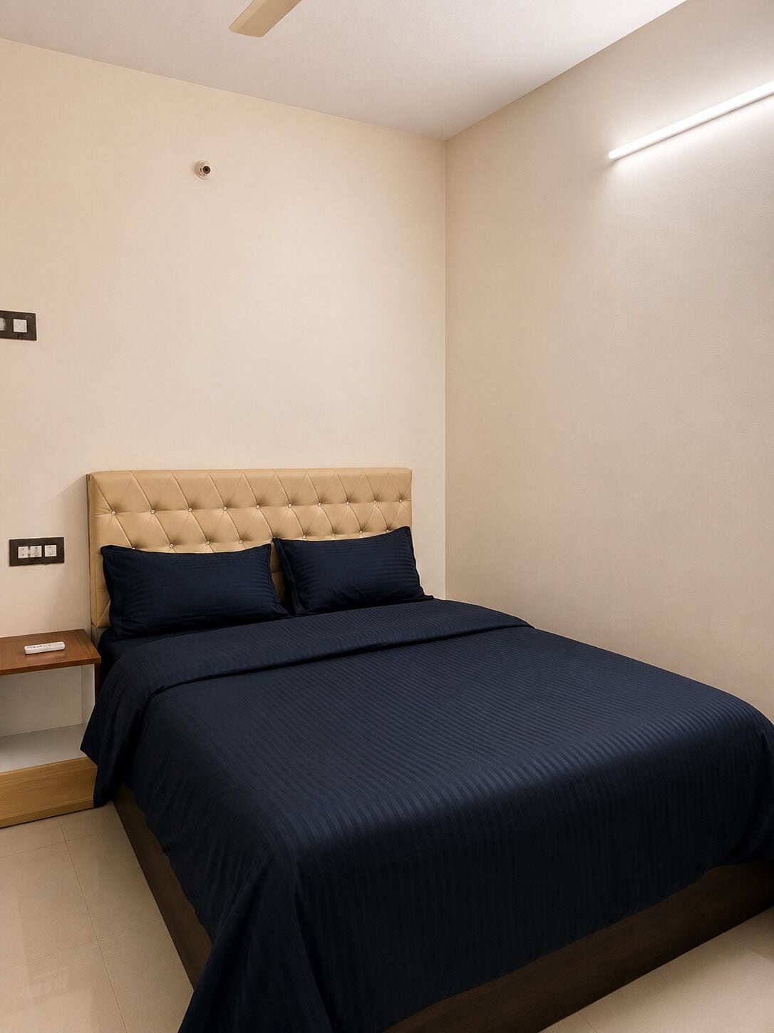 Quarto com cabeceira estofada, edredom escuro e mesa de cabeceira com controle remoto.
