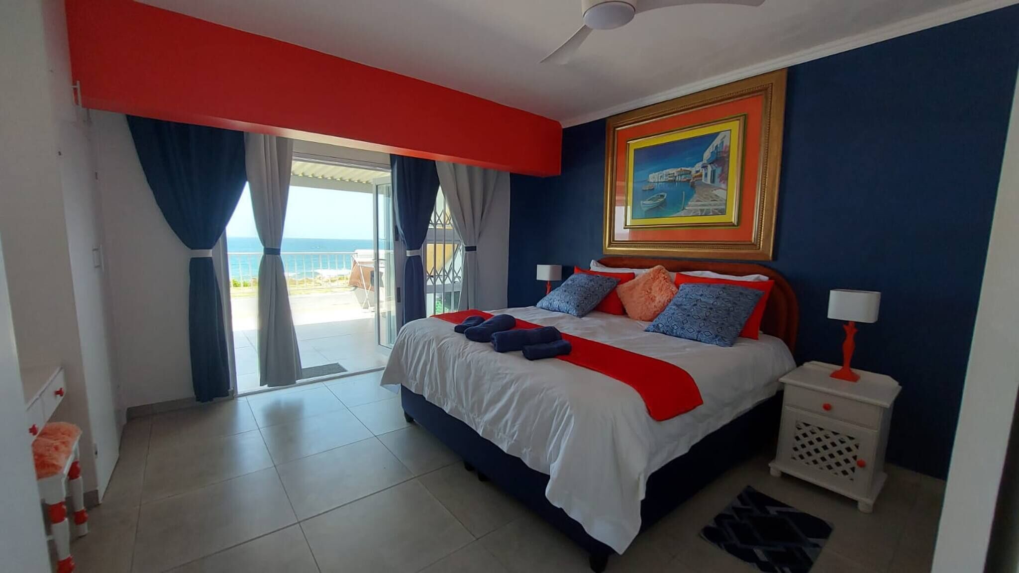 Um quarto com cama, mesa de cabeceira, luminária, pintura e vista para o mar pela janela.