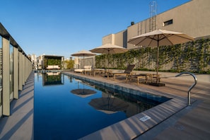 Piscina no terraço com espreguiçadeiras e guarda-sóis, rodeada por uma fachada de prédio moderno.