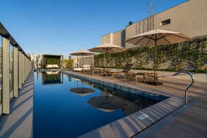 Piscina no terraço com espreguiçadeiras e guarda-sóis, rodeada por uma fachada de prédio moderno.