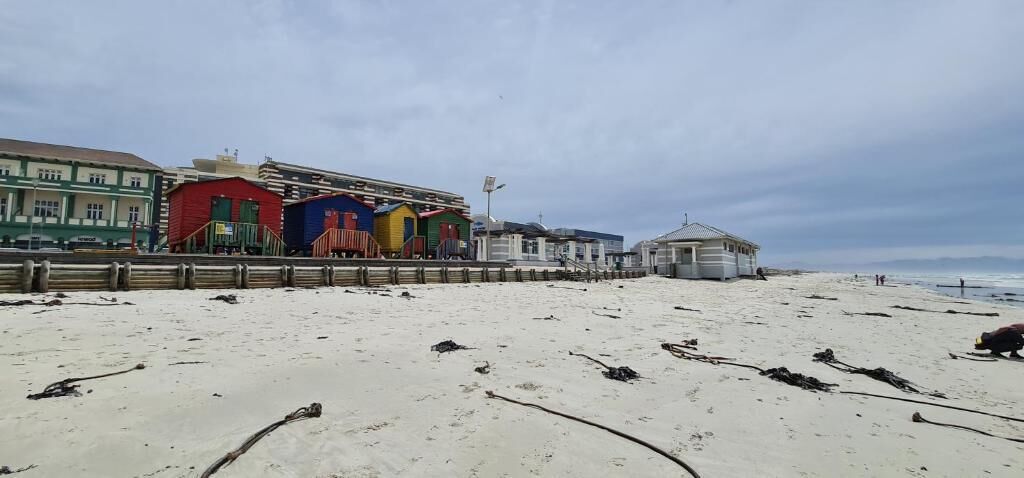 Uma praia com uma fileira de edifícios coloridos, margem arenosa e uma pequena construção à direita.