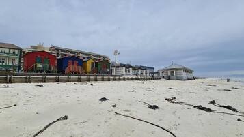 Uma praia com uma fileira de edifícios coloridos, margem arenosa e uma pequena construção à direita.