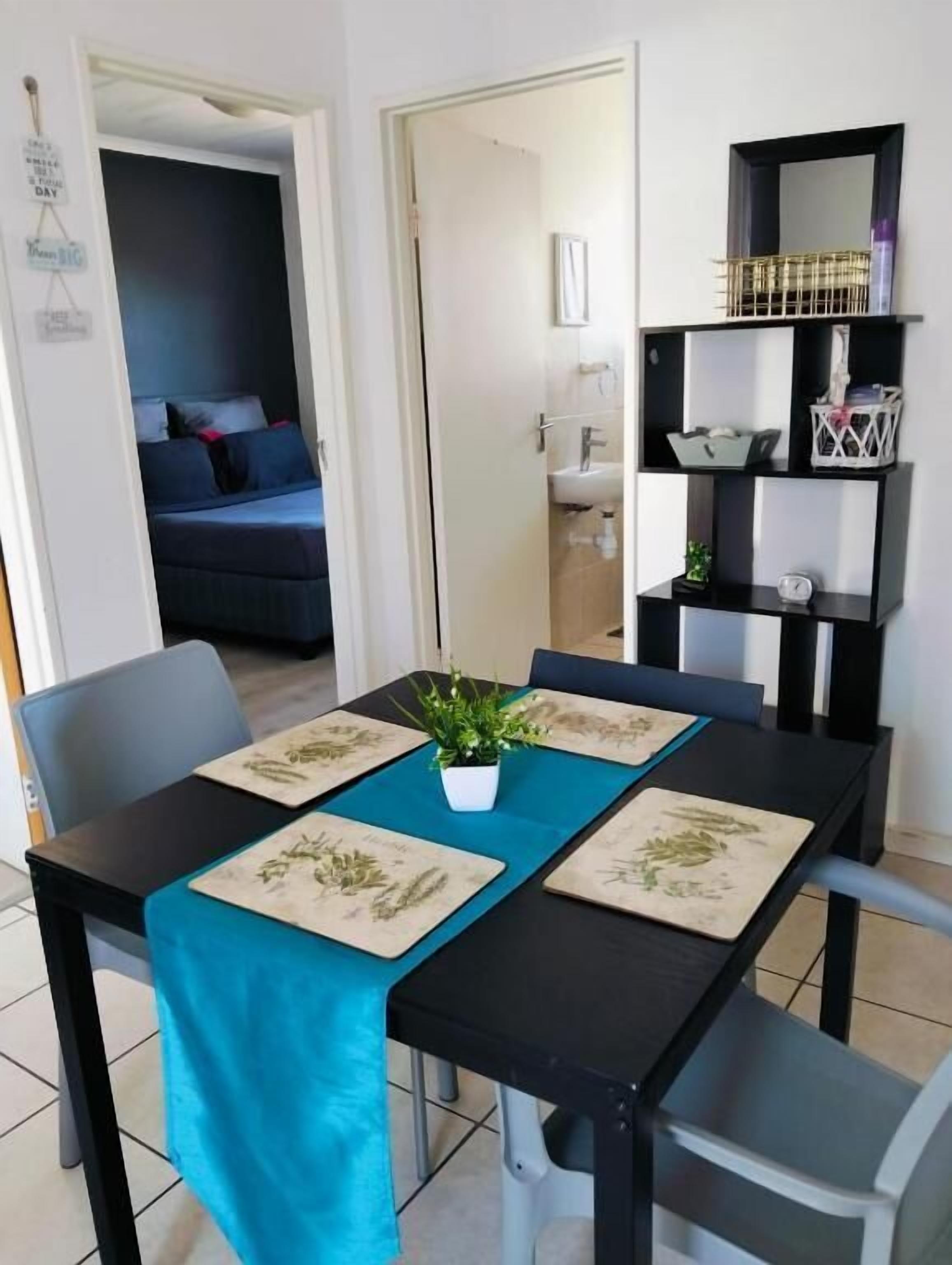 Uma área de refeições com uma mesa posta para quatro pessoas, uma planta em vaso e uma prateleira preta fixada na parede com diversos objetos.