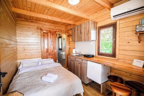 Interior de uma cabana de madeira com cama, cozinha compacta e um pequeno frigorífico.