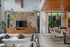 Uma sala de estar moderna com uma parede de pedra, televisão de tela plana, unidade de entretenimento de madeira, sofá e um espaço de refeições com mesa e cadeiras.