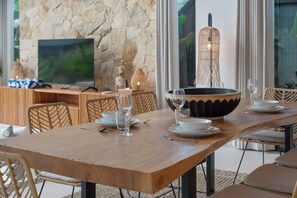 Uma área de refeições com uma mesa de madeira posta para dois, uma parede de pedra e uma televisão ao fundo.