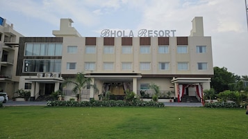 Um hotel chamado 'BHOLA RESORT', com design arquitetônico moderno, com vários andares, entrada central com detalhes decorativos e um gramado bem cuidado em primeiro plano.