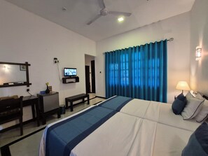 Quarto de hotel com cama, escrivaninha, televisão, ventoinha no teto e janela com cortinas.