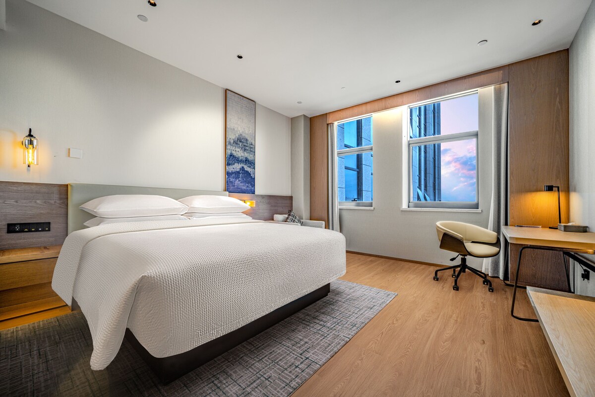 Quarto de hotel moderno com uma cama grande, uma escrivaninha e janela com vista para a cidade.