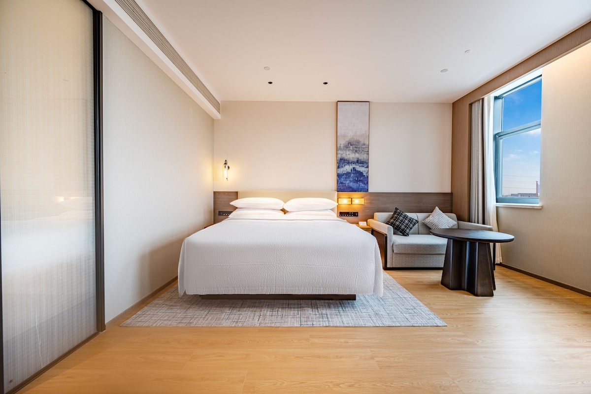 Quarto de hotel moderno com uma cama grande, área de estar com uma mesa e uma janela ampla com vista.