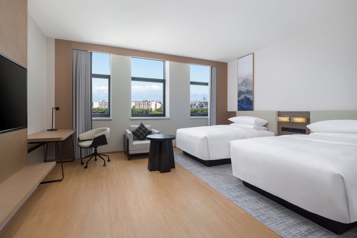 Quarto de hotel com duas camas, uma escrivaninha, uma cadeira e vista para a paisagem urbana.