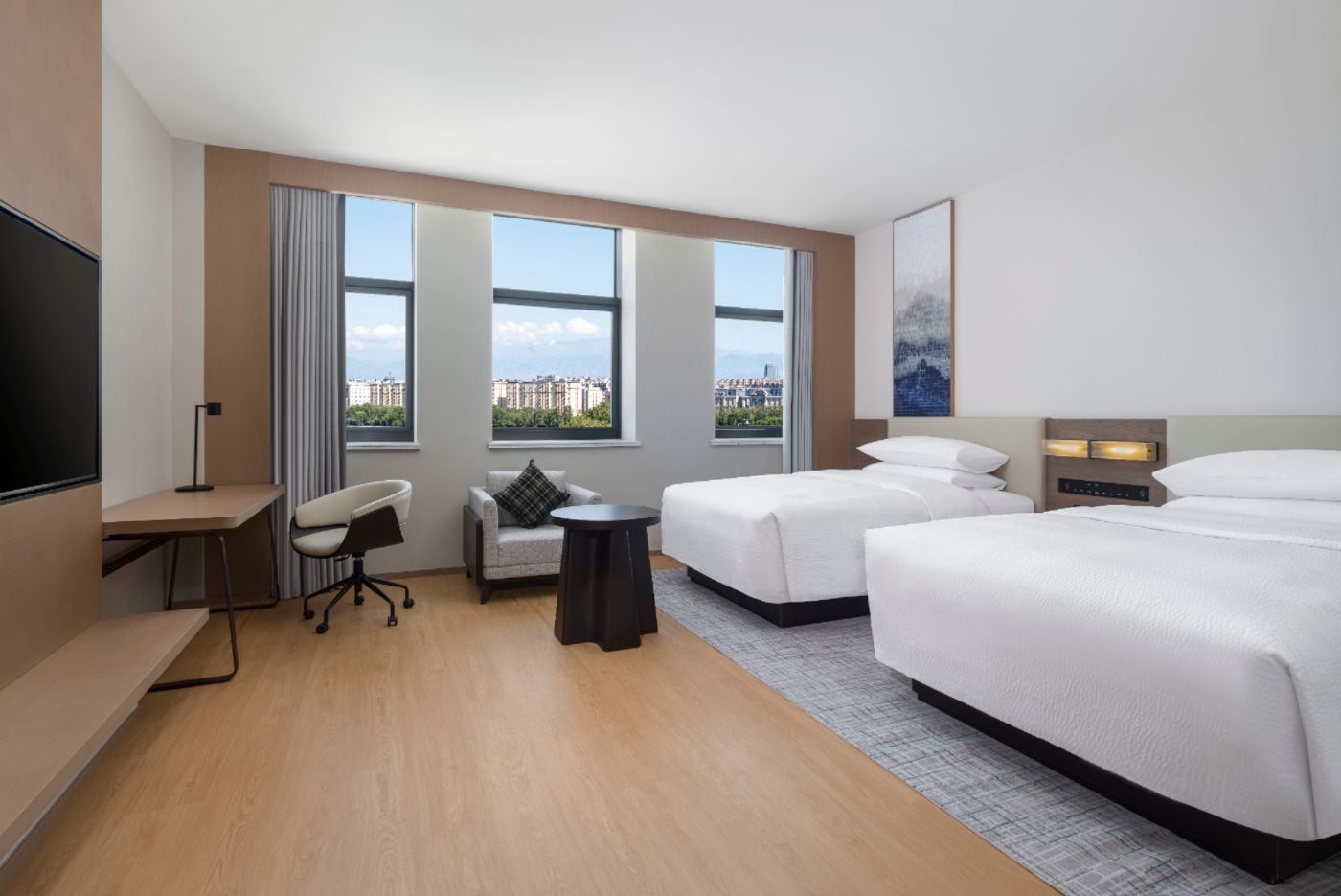 Quarto de hotel com duas camas, escrivaninha, cadeira e janelas amplas com vista para a cidade.