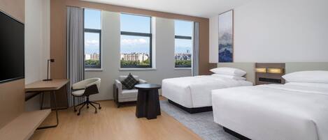 Quarto de hotel com duas camas, escrivaninha, cadeira e janelas amplas com vista para a cidade.