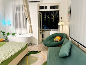 Quarto moderno com sofá verde, cama com roupa de cama listrada, banheira e janela com luzes de cordão.