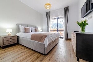 Quarto com cama, mesa de cabeceira, luminária, cômoda, televisão e janela com cortinas.