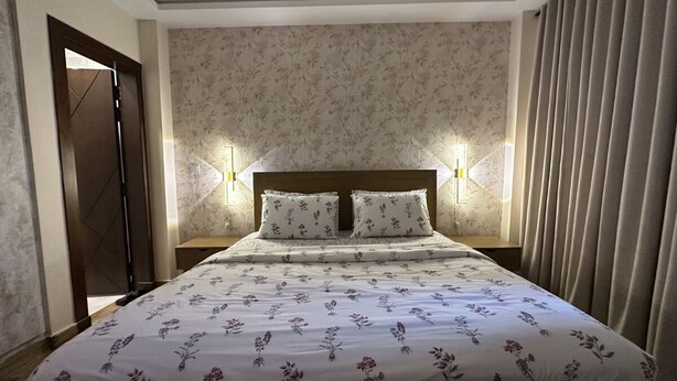 Quarto de hotel com cama, roupa de cama estampada com flores, duas luminárias de cabeceira e uma porta.