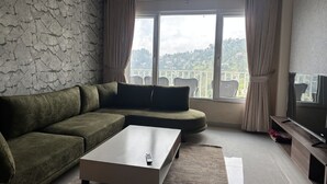 Uma sala de estar com um sofá verde, uma mesa de centro branca, uma TV de tela plana e uma grande janela com cortinas.