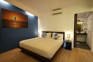 Quarto de hotel com cama, pintura de pôr do sol, ar condicionado, banheiro com espelho e mesa de cabeceira com luminária.