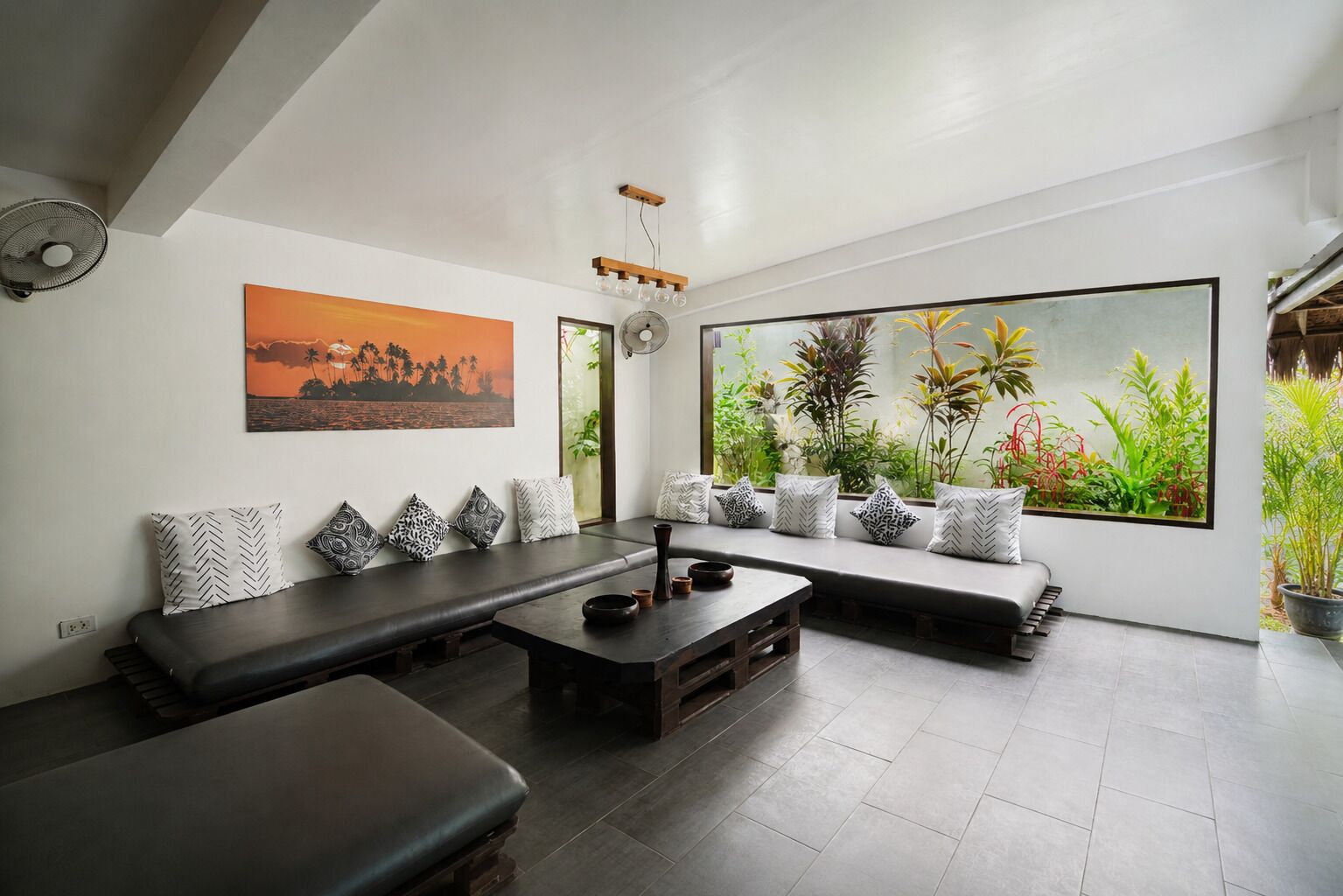 Uma sala de estar moderna com um sofá, um banco e uma mesa de jantar. Há grandes janelas com vista para plantas e uma pintura do pôr do sol na parede.