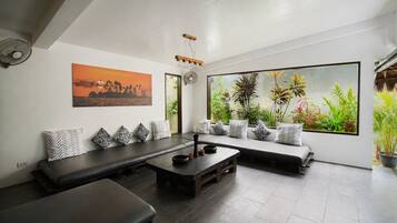 Uma sala de estar moderna com um sofá, um banco e uma mesa de jantar. Há grandes janelas com vista para plantas e uma pintura do pôr do sol na parede.
