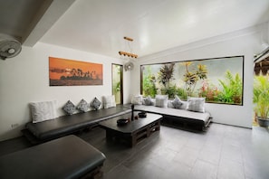 Uma sala de estar moderna com um sofá, um banco e uma mesa de jantar. Há grandes janelas com vista para plantas e uma pintura do pôr do sol na parede.