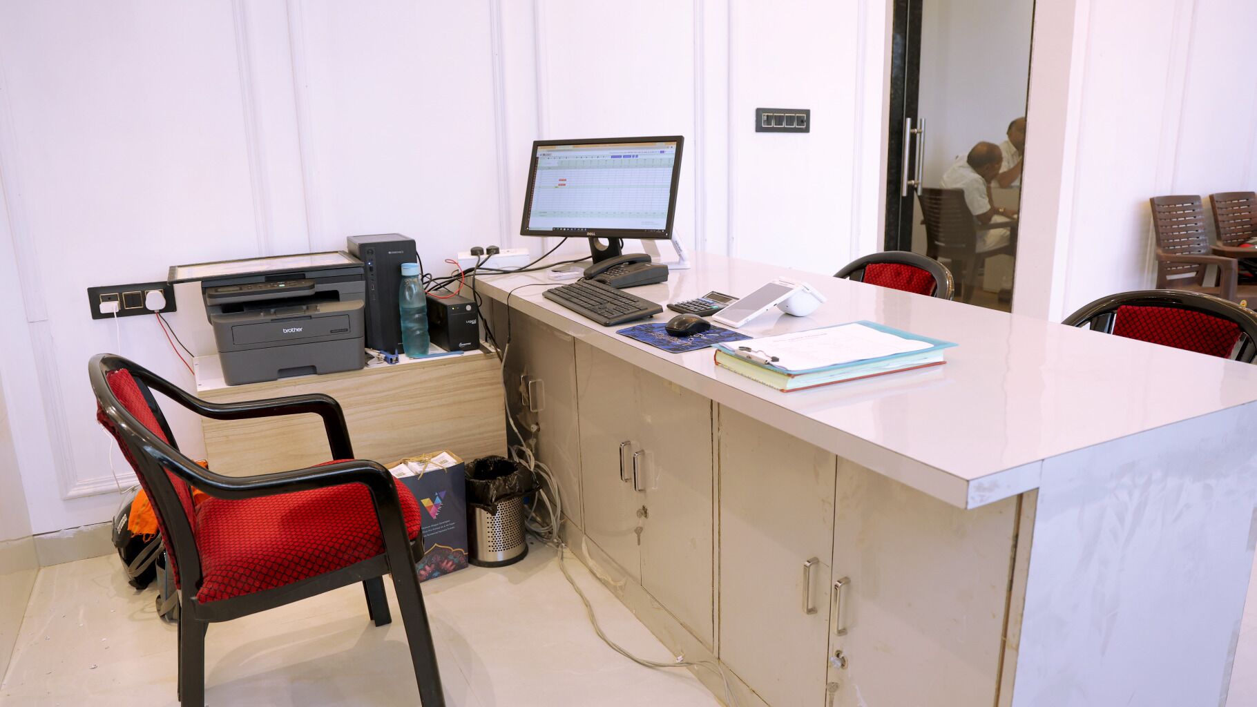 Balcão de recepção com computador, impressora e telefone. Há duas cadeiras e uma lixeira.