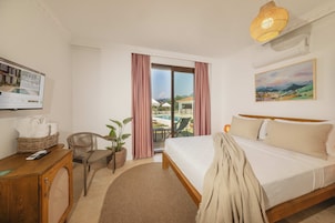 Quarto de hotel com cama, escrivaninha com computador, cadeira, planta e um quadro na parede.