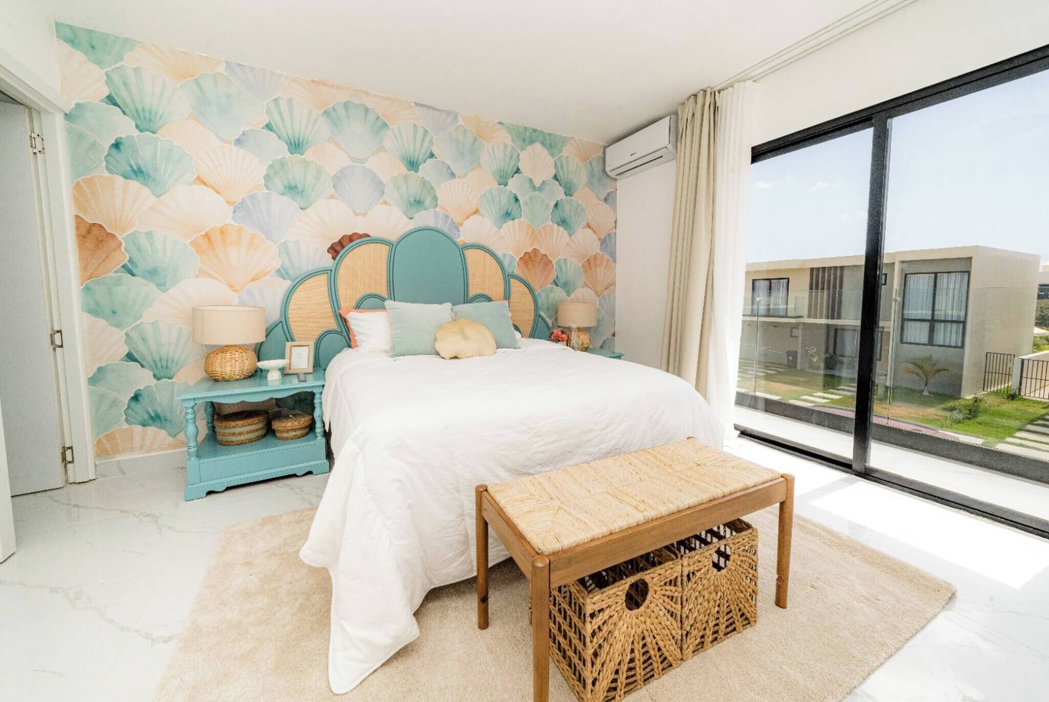 Quarto com uma cama grande, uma mesinha de cabeceira, um banquinho de vime e vista para um edifício através de uma janela ampla.