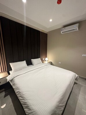 Quarto de hotel com uma cama grande, uma mesa de cabeceira com abajur, um aparelho de ar condicionado e um ponto de luz fixado na parede.