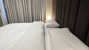 Quarto de hotel com duas camas, uma mesa de cabeceira, uma luminária e uma janela com cortinas.