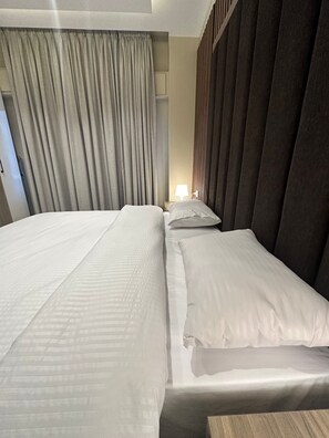 Quarto de hotel com duas camas, uma mesa de cabeceira, uma luminária e uma janela com cortinas.
