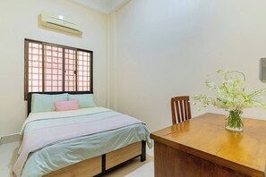 Quarto com uma cama, uma mesa de madeira, uma cadeira e uma janela com padrão quadriculado.