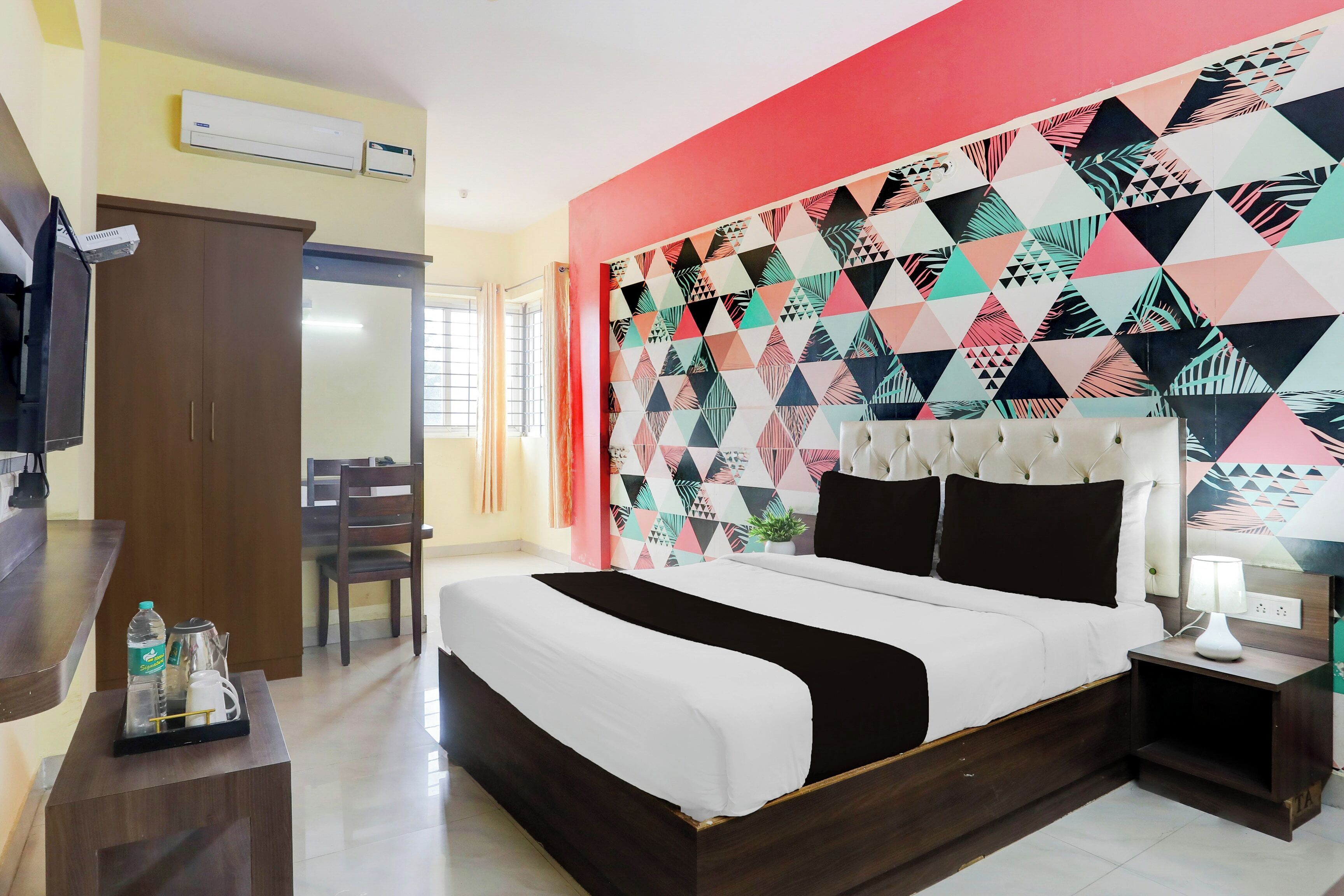 Quarto de hotel com cama, escrivaninha, cadeira, televisão e parede com padrão geométrico.