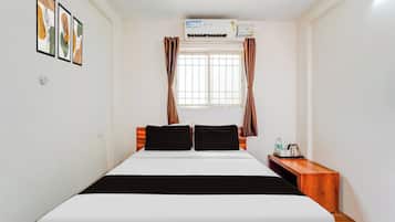 Quarto de hotel com uma cama, janela com cortinas, ar condicionado e uma pequena mesa de madeira com chaleira e chávenas.