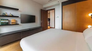 Quarto de hotel com cama, televisão fixada na parede, guarda-roupa de madeira e uma estante com livros.
