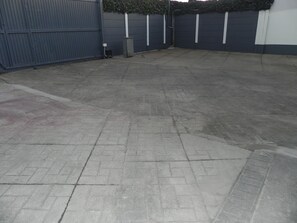 Uma área externa pavimentada com um portão azul, uma lixeira e uma parede com linhas verticais.
